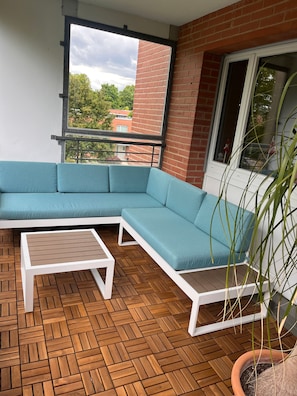 Uma varanda com um sofá azul em formato de L, uma mesa de madeira e um vaso com planta.