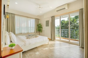 Um quarto com uma cama, uma mesa de cabeceira com uma planta, uma janela grande com cortinas e uma varanda com vista para o verde.