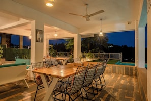 Uma área de refeições com mesa de madeira e cadeiras de metal, grandes janelas e um ventilador de teto.