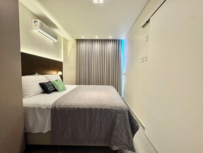 Quarto de hotel estreito com uma cama, cabeceira, janela com cortinas e um ar condicionado fixado na parede.