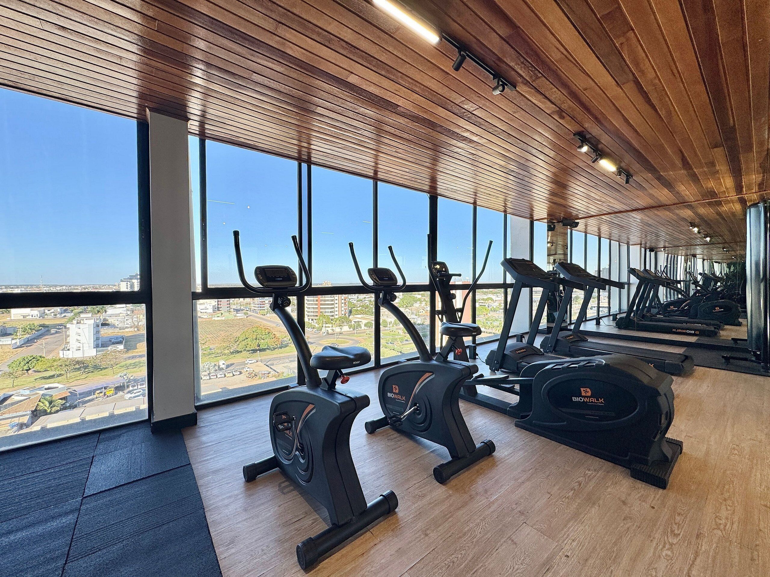 Uma academia com equipamentos de cardio, amplas janelas e vista para a cidade.