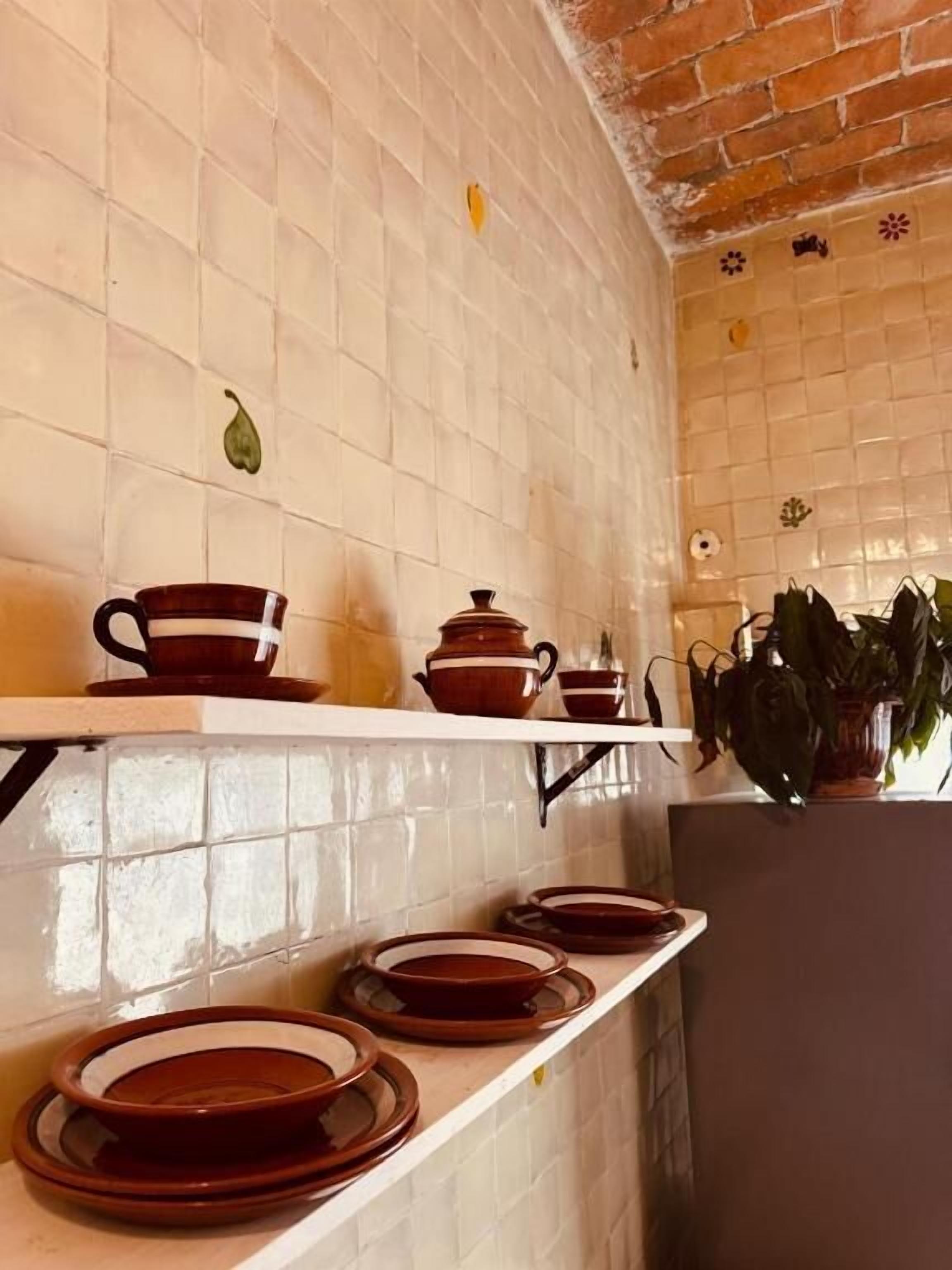 Uma cozinha com paredes revestidas de azulejos brancos, prateleiras com louças marrons e um vaso de planta.