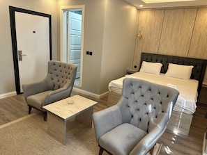 Quarto de hotel com cama, duas poltronas, uma mesinha e chão de madeira.