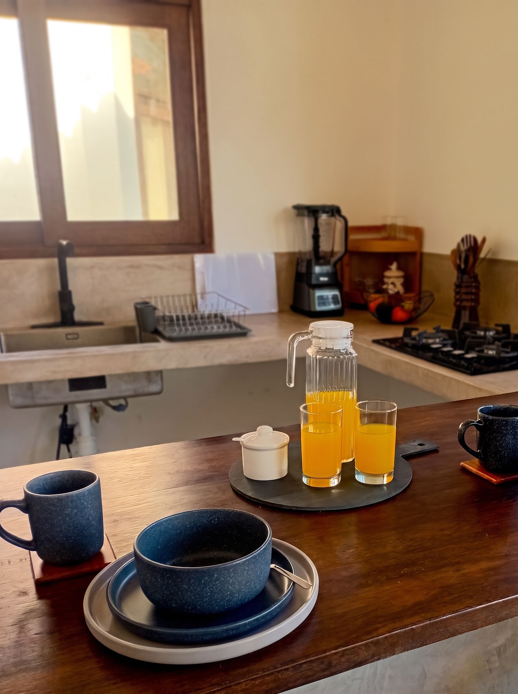 Uma bancada de cozinha com uma cafeteira, um bule e uma torradeira. Há também duas chávenas e um prato com uma tigela.