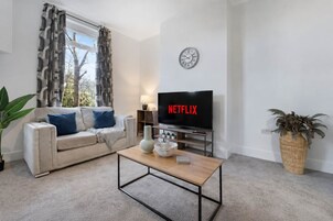 Uma sala de estar com sofá, mesa de centro, televisão, relógio e janela com cortinas.