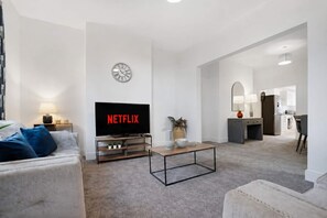 Uma sala de estar moderna com uma televisão de tela plana exibindo 'NETFLIX', um relógio na parede e uma área de cozinha ao fundo.