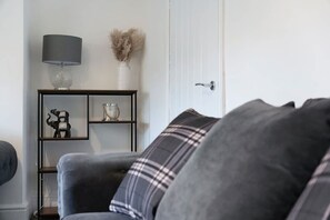 Uma sala de estar com um sofá cinzento, uma mesa lateral com abajur e objetos decorativos, e uma porta com puxador.