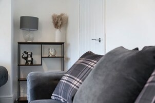 Uma sala de estar com um sofá cinzento, uma mesa lateral com abajur e objetos decorativos, e uma porta com puxador.