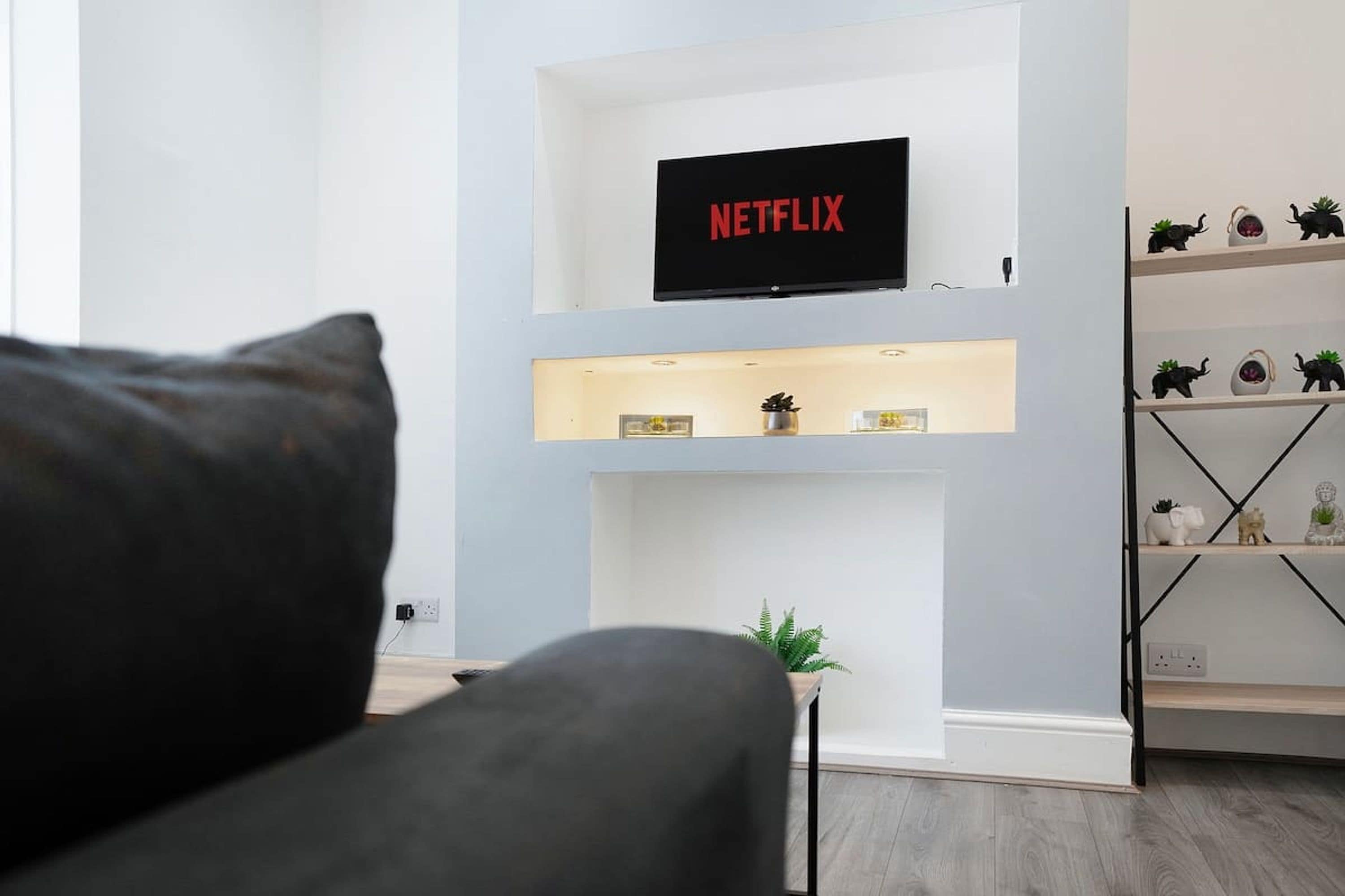 Uma sala de estar com uma televisão montada exibindo 'NETFLIX' em letras vermelhas, uma lareira com objetos decorativos e uma estante com pequenas figuras.