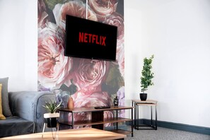 Sala de estar com televisão de tela plana fixada em papel de parede floral, um sofá cinza, uma mesinha de centro de madeira, um vaso de planta e uma mesa lateral com controle remoto.