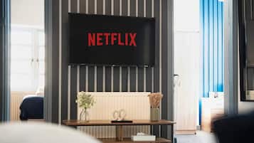 Quarto com televisão na parede exibindo 'NETFLIX' em letras vermelhas, mesa consola de madeira com um vaso e tesouras, e vista para uma cortina azul e uma porta.
