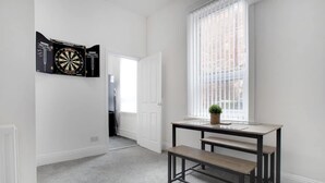 Um quarto com um tabuleiro de dardos, uma janela com persianas, um banco e uma pequena mesa com uma planta.