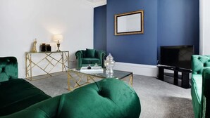 Uma sala de estar com uma parede azul de destaque, uma televisão de tela plana grande e móveis estofados verdes.
