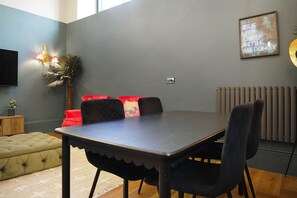 Quarto moderno com parede cinzenta, mesa preta e cadeiras vermelhas e pretas. Há uma televisão à esquerda e um radiador à direita.
