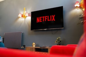 Uma sala de estar moderna com uma televisão de tela plana grande fixada na parede exibindo 'NETFLIX' em letras vermelhas, um radiador, uma mesa pequena com uma decoração de caveira e uma planta em vaso.