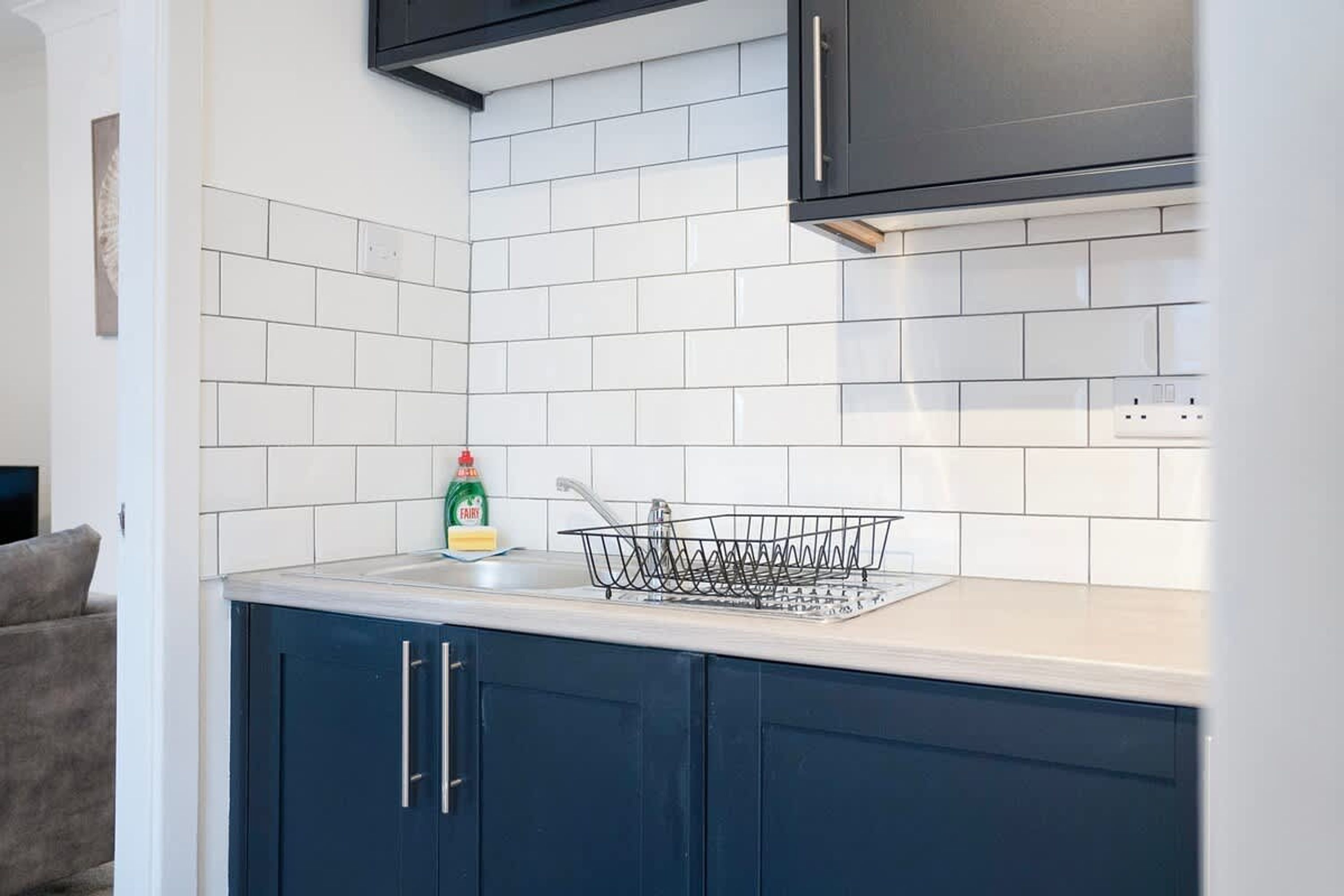 Cozinha com painel de azulejos brancos, armários azul escuro, pia de aço inoxidável e escorredor de louça.
