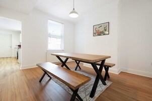 Uma sala de jantar com mesa e banco de madeira, luminária pendente e janela com persianas.