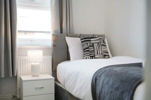 Uma cama bem arrumada com edredom branco e almofada estampada em preto e branco, uma mesa de cabeceira branca com luminária, e cortinas cinzas numa janela.