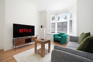 Uma sala de estar com uma televisão exibindo 'NETFLIX', uma mesa de centro de madeira, uma poltrona azul-turquesa e um sofá cinza.