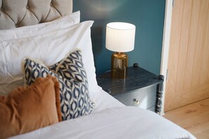 Uma cama bem arrumada com roupa de cama branca e almofadas estampadas, uma mesa de cabeceira com abajur e uma parede de cor escura.