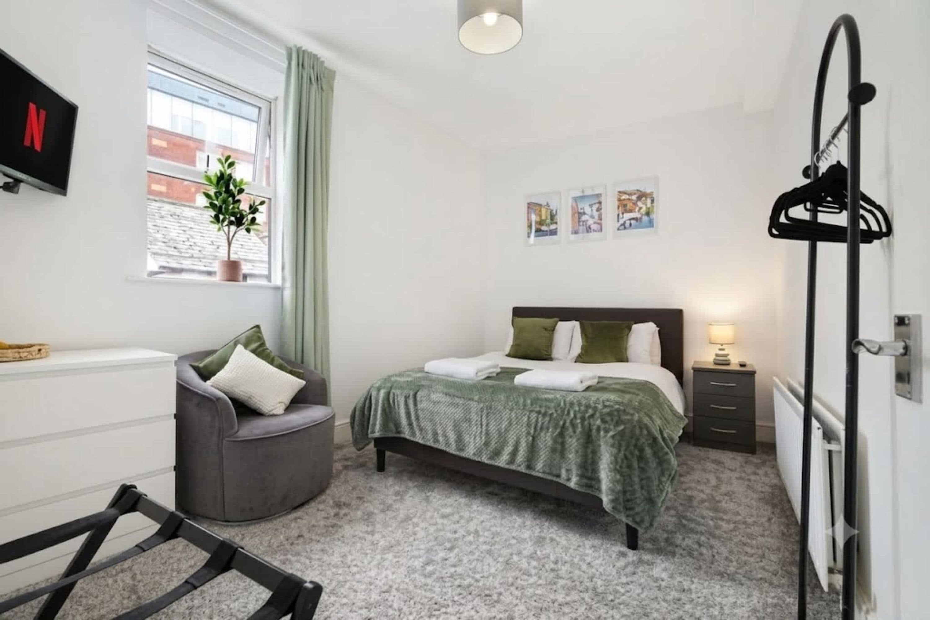 Quarto com cama, escrivaninha, cadeira, radiador, televisão e janela com uma planta.