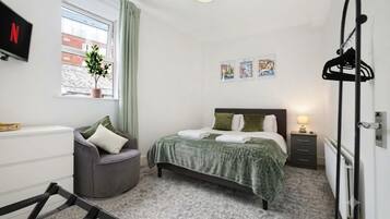 Quarto com cama, escrivaninha, cadeira, radiador, televisão e janela com uma planta.