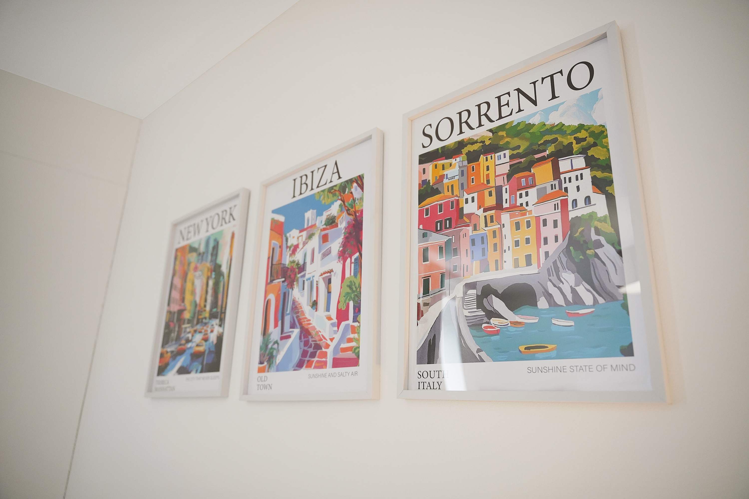 Três obras de arte emolduradas com os títulos 'NEW YORK', 'IBIZA' e 'Sorrento' numa parede branca.