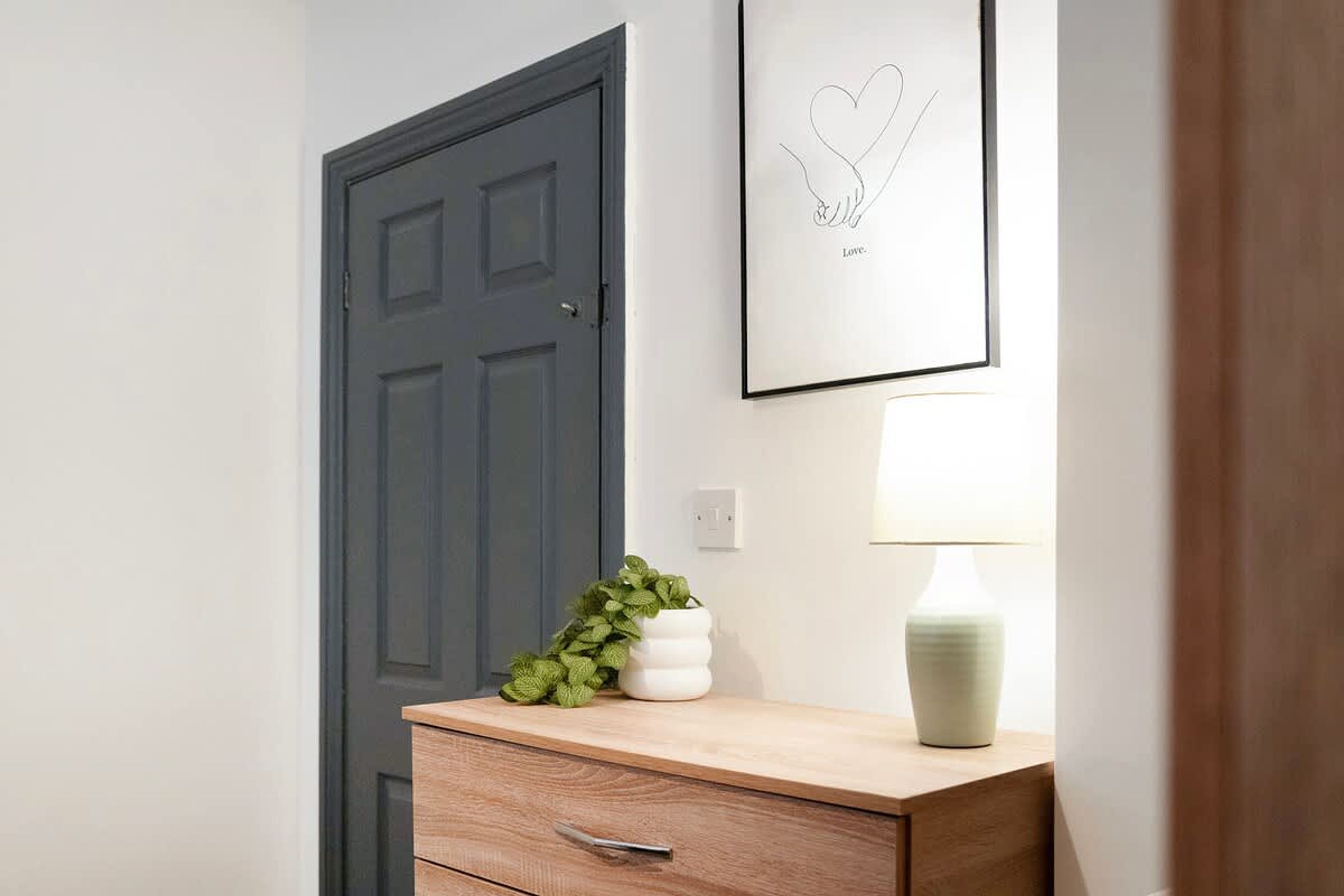 Um corredor com uma porta escura, um quadro com o desenho de um coração, uma planta em vaso, um vaso branco e uma luminária sobre um balcão de madeira.