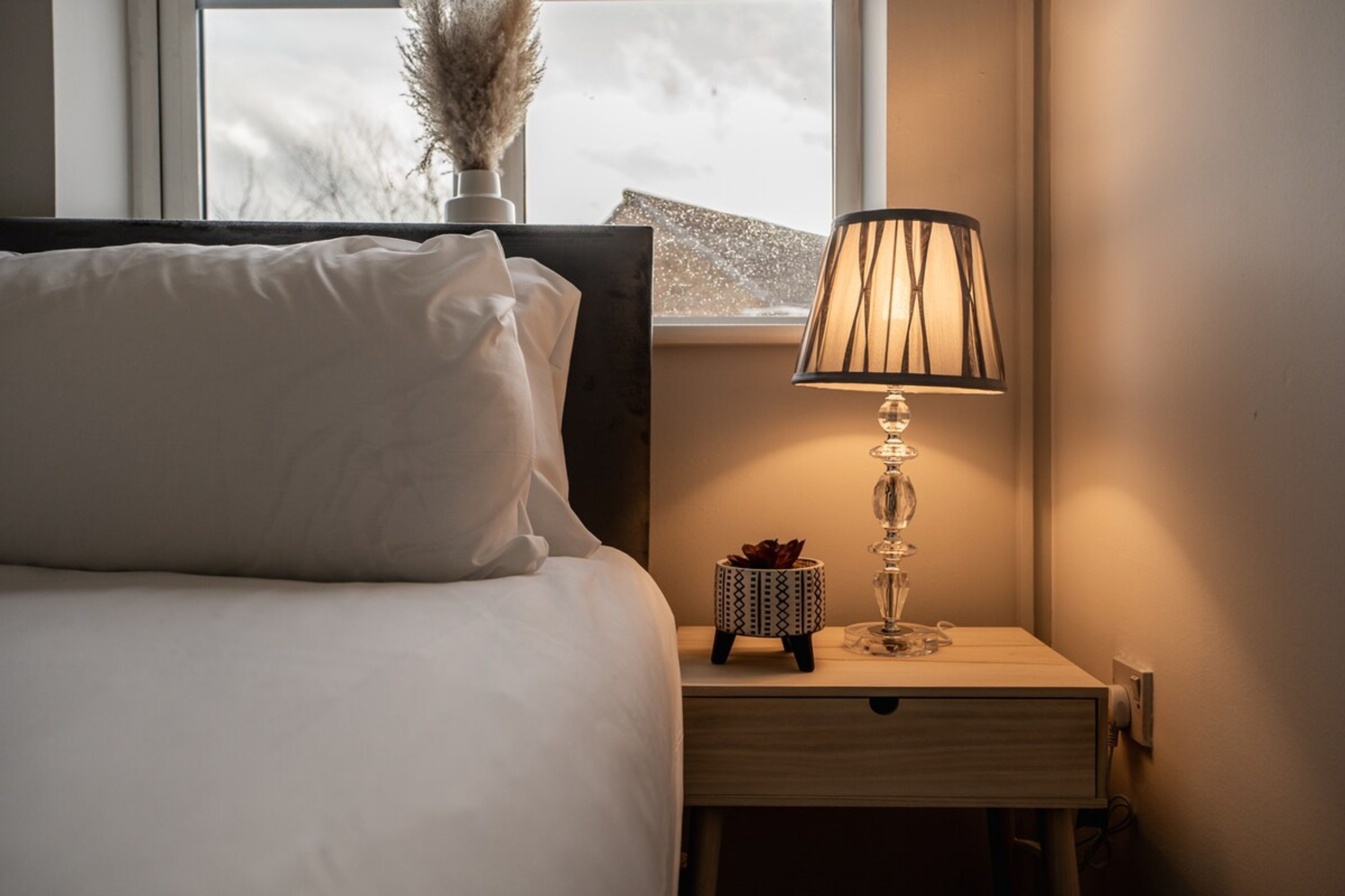 Uma cama bem arrumada com lençóis brancos, um abajur, uma pequena planta decorativa e uma janela com vista para uma paisagem nevada.