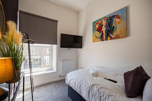 Quarto com cama, televisão fixada na parede, janela com vista para edifícios e um quadro na parede.