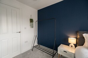 Quarto com porta branca, parede azul, suporte de metal preto, luminária de cabeceira e pequena mesa de apoio.
