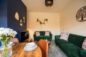 Uma sala de estar com um sofá verde, uma mesa de jantar de madeira com pratos brancos e um vaso de flores brancas.