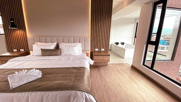 Quarto de hotel moderno com uma cama grande, cabeceira de madeira, mesa de cabeceira e vista para a cidade através de uma janela ampla.
