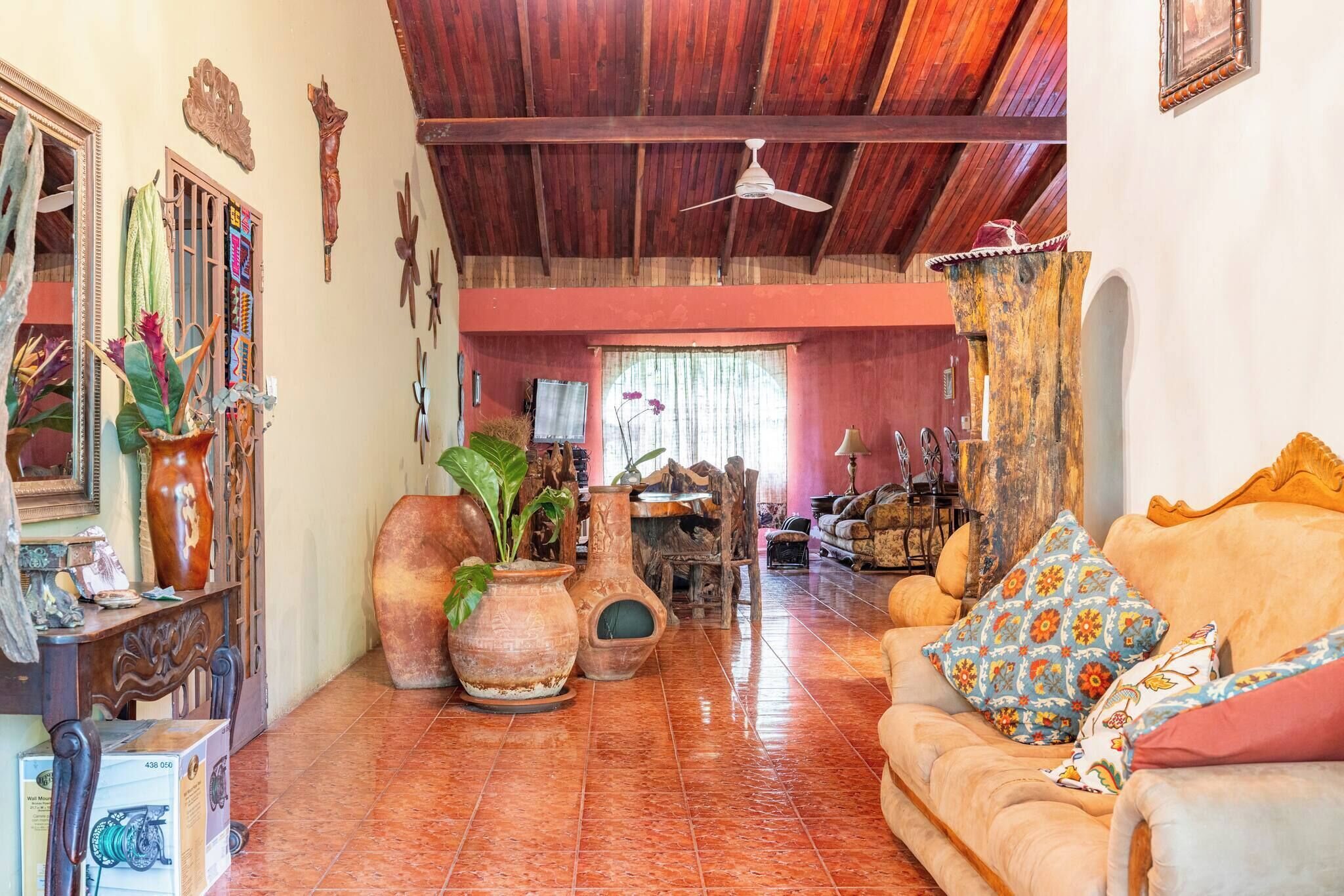 Uma sala de estar ampla com teto de madeira, um sofá com almofadas coloridas e diversos objetos decorativos, incluindo um vaso grande e uma escultura de madeira.