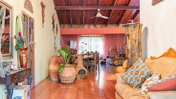 Uma sala de estar ampla com teto de madeira, um sofá com almofadas coloridas e diversos objetos decorativos, incluindo um vaso grande e uma escultura de madeira.