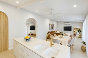 Uma cozinha moderna com ilha central, área de refeições com mesa redonda e sala de estar com sofá e televisão.