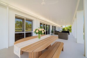Uma ampla área de refeições ao ar livre, com mesa e bancos de madeira, rodeada por grandes janelas com vista para o verde.