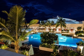 Vista noturna de um hotel com piscina, palmeiras e um edifício moderno com cúpula.