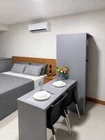 Quarto de hotel compacto com uma cama, uma pequena mesa de jantar com cadeiras, um vaso com flores e um ar condicionado.