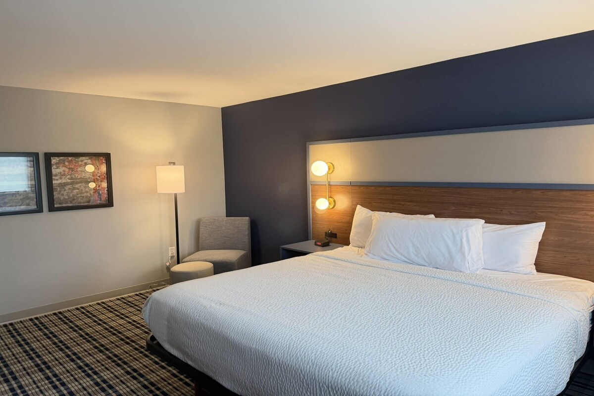 Quarto de hotel com uma cama grande, uma cadeira, mesa de cabeceira e duas luminárias na parede.