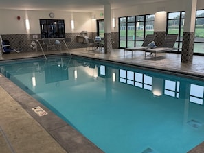 Piscina com espreguiçadeiras e uma mesa, cercada por janelas e um painel de azulejos.