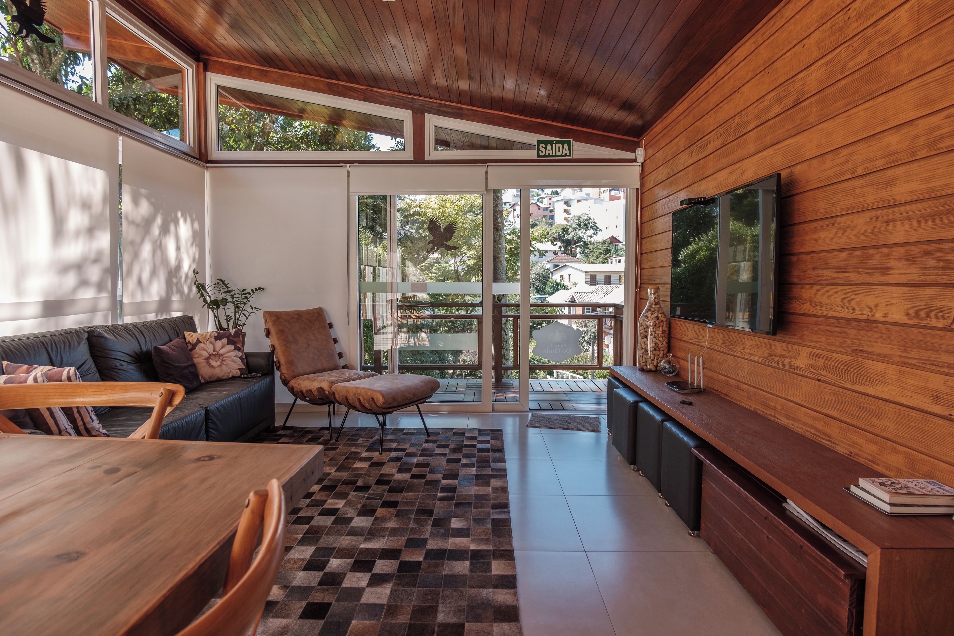 Interior moderno com teto de madeira, um sofá, uma poltrona, uma mesa de jantar e uma televisão na parede.