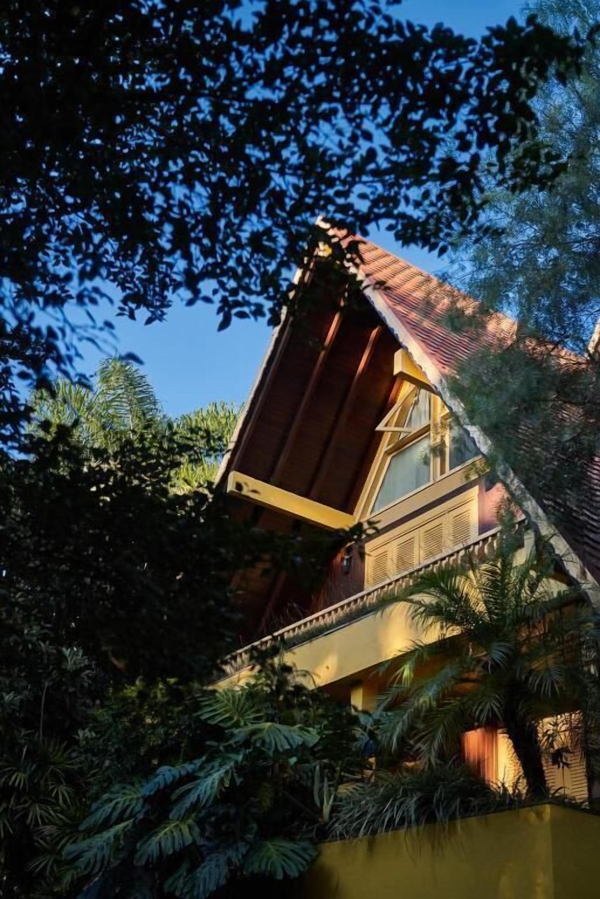 Um edifício com telhado inclinado, rodeado por vegetação densa e um corpo de água.