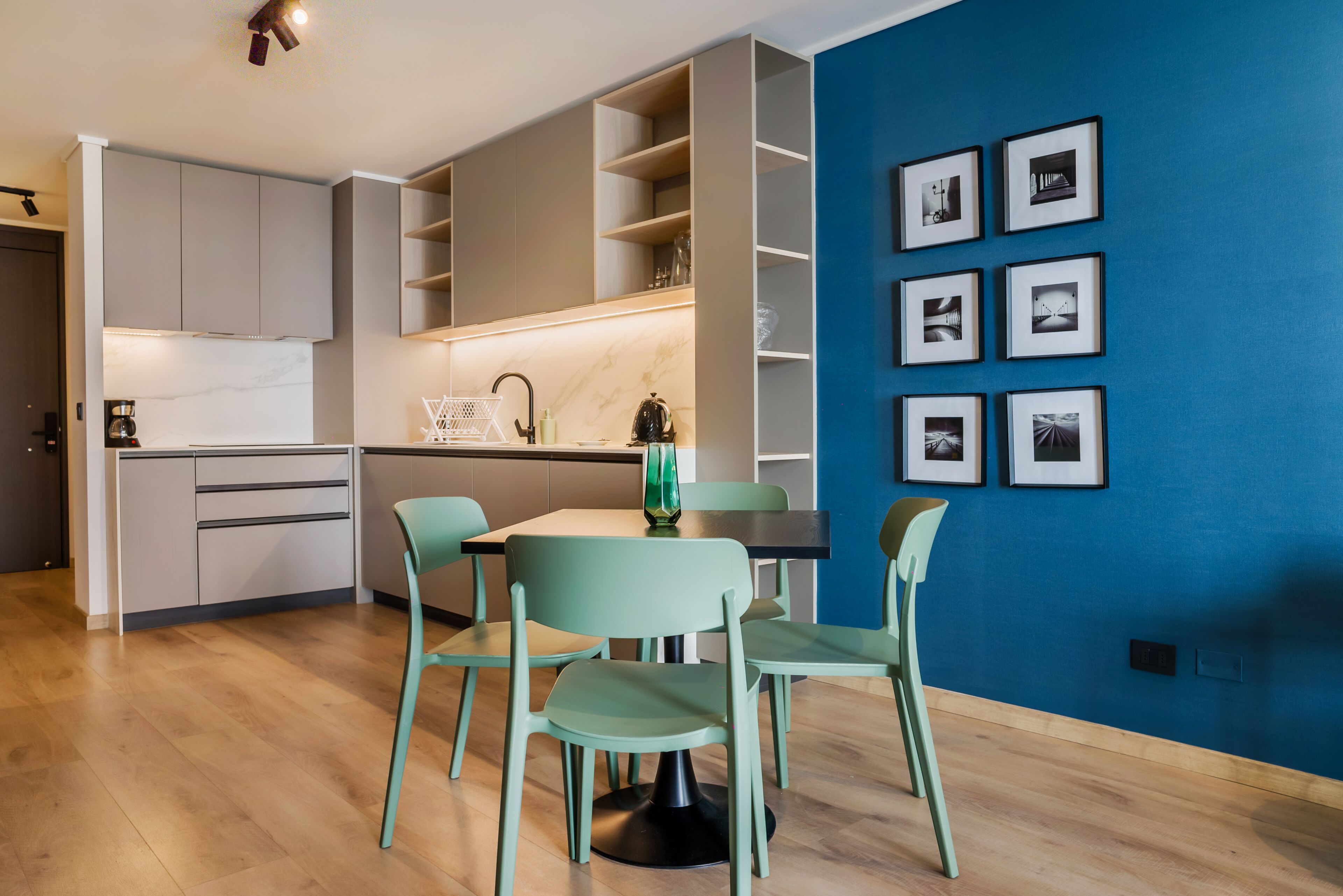 Uma cozinha moderna com armários embutidos, um espaço de refeições com cadeiras cor turquesa e uma parede azul decorada com quadros.