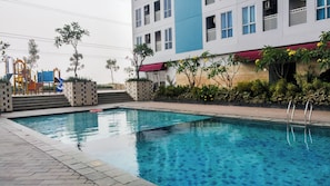 Piscina com superfície de água azul cristalina, ladeada por um calçadão pavimentado. Escadas levam a um edifício com fachada azul e branca, e um parquinho com equipamentos coloridos é visível ao fundo.