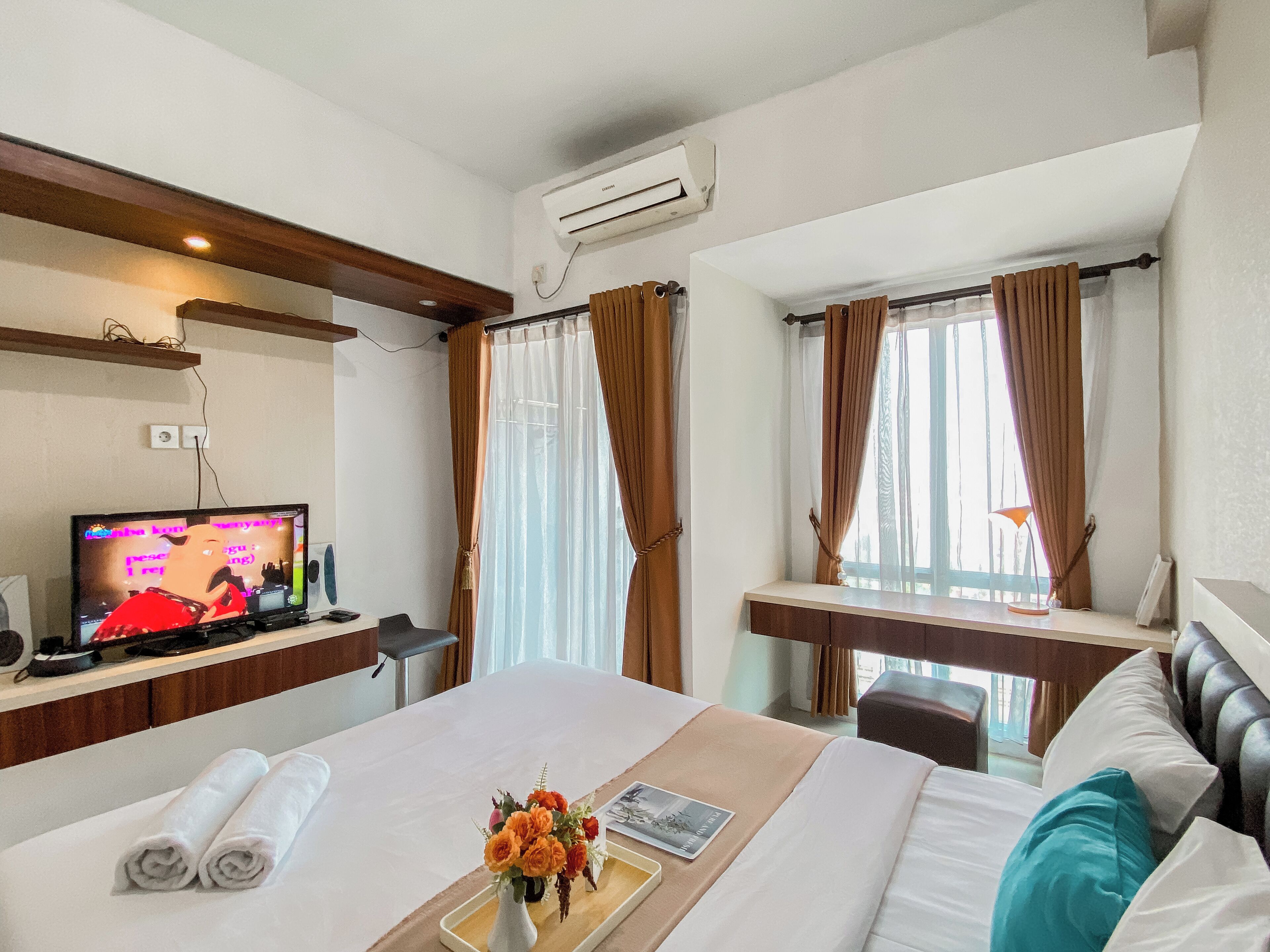 Quarto de hotel com cama, televisão, escrivaninha e janela com cortinas.
