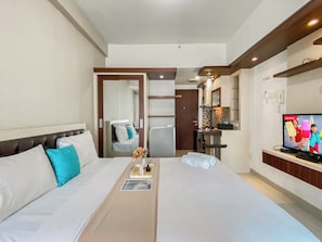 Quarto de hotel com cama, televisão, pequeno kitchenette e banheiro visível por uma porta.