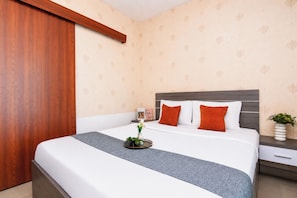 Cama bem arrumada com roupa de cama branca e cinza, dois travesseiros laranja, uma pequena planta na mesa de cabeceira e uma cabeceira de madeira.