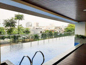 Piscina interna com paredes de vidro transparente, deck de madeira e vista para edifícios e áreas verdes ao redor.