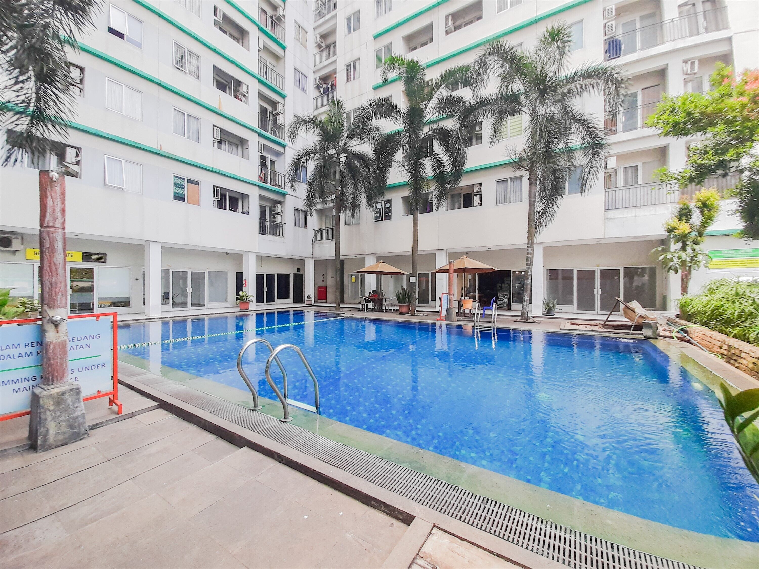 Piscina rodeada por um edifício com vários andares, varandas, palmeiras e áreas de estar ao ar livre.